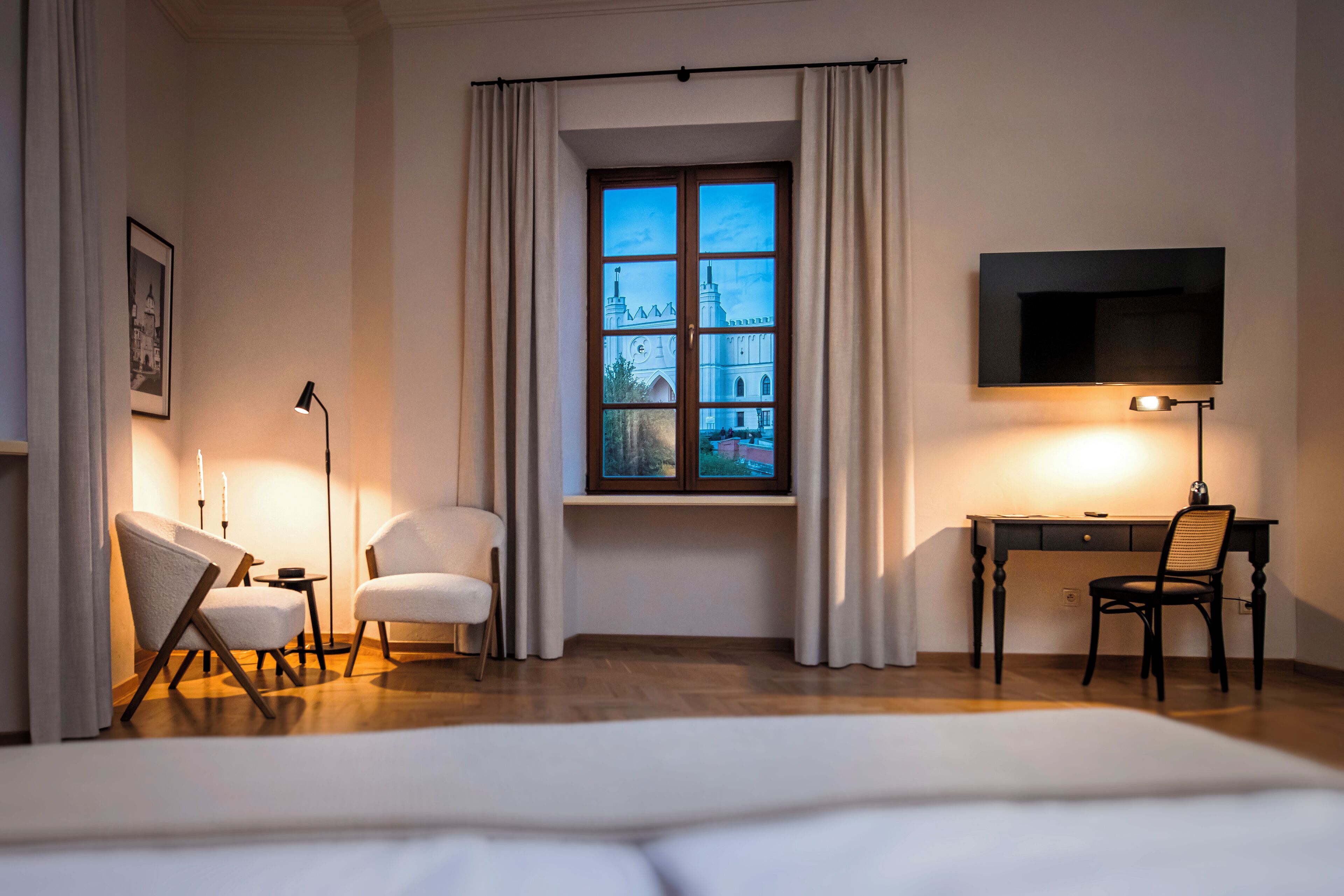 Quarto de hotel com uma cama grande, uma escrivaninha com cadeira, dois poltronas e uma televisão presa na parede, além de uma janela com cortinas.