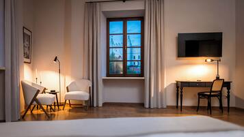 Quarto de hotel com uma cama grande, uma escrivaninha com cadeira, dois poltronas e uma televisão presa na parede, além de uma janela com cortinas.
