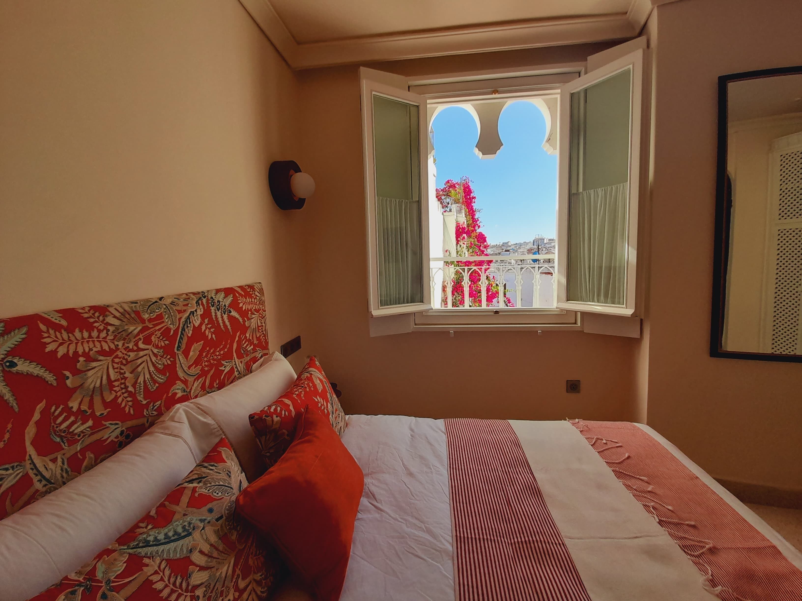 Quarto com cama, janela com vista para uma varanda e luminária fixada na parede.