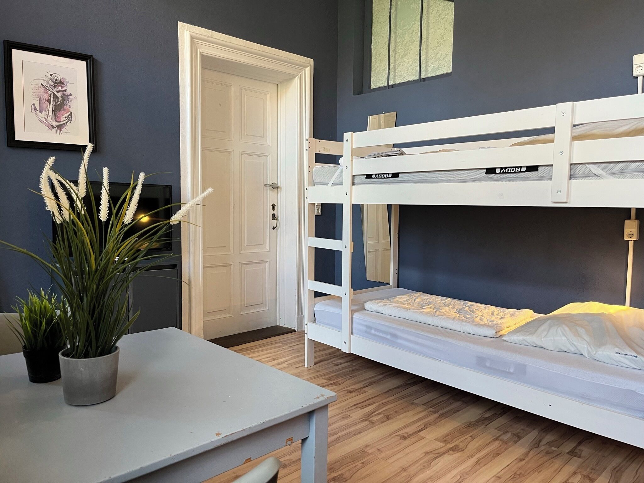 Uma cama beliche com colchão e travesseiros, uma escrivaninha branca com uma planta, um quadro na parede e uma porta.