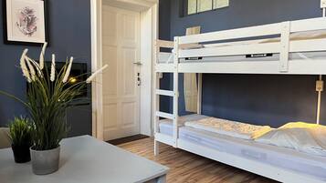 Uma cama beliche com colchão e travesseiros, uma escrivaninha branca com uma planta, um quadro na parede e uma porta.
