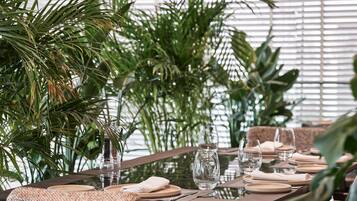 Mesa de jantar posta com pratos, copos e talheres, rodeada por plantas verdes exuberantes.