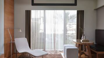 Quarto de hotel moderno com uma janela grande, uma luminária de pé, uma poltrona estofada, uma mesa redonda pequena e um sofá.