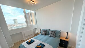 Quarto moderno com uma cama grande, uma mesa de cabeceira, abajur e vista para um horizonte urbano através de amplas janelas.