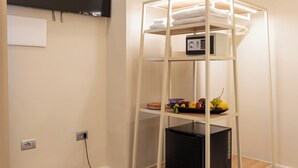 Quarto com televisão integrada, prateleira branca com caixa de armazenamento preta e piso de madeira.