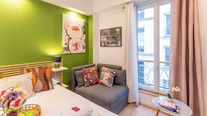 Quarto de hotel com uma cama, um sofá cinzento, uma mesinha com um vaso, e uma janela com cortinas.