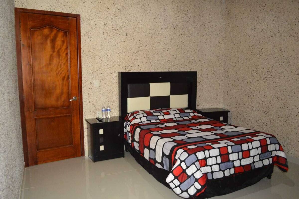 Um quarto com porta de madeira, uma cama com cabeceira xadrez, duas mesas de cabeceira e um edredom estampado.