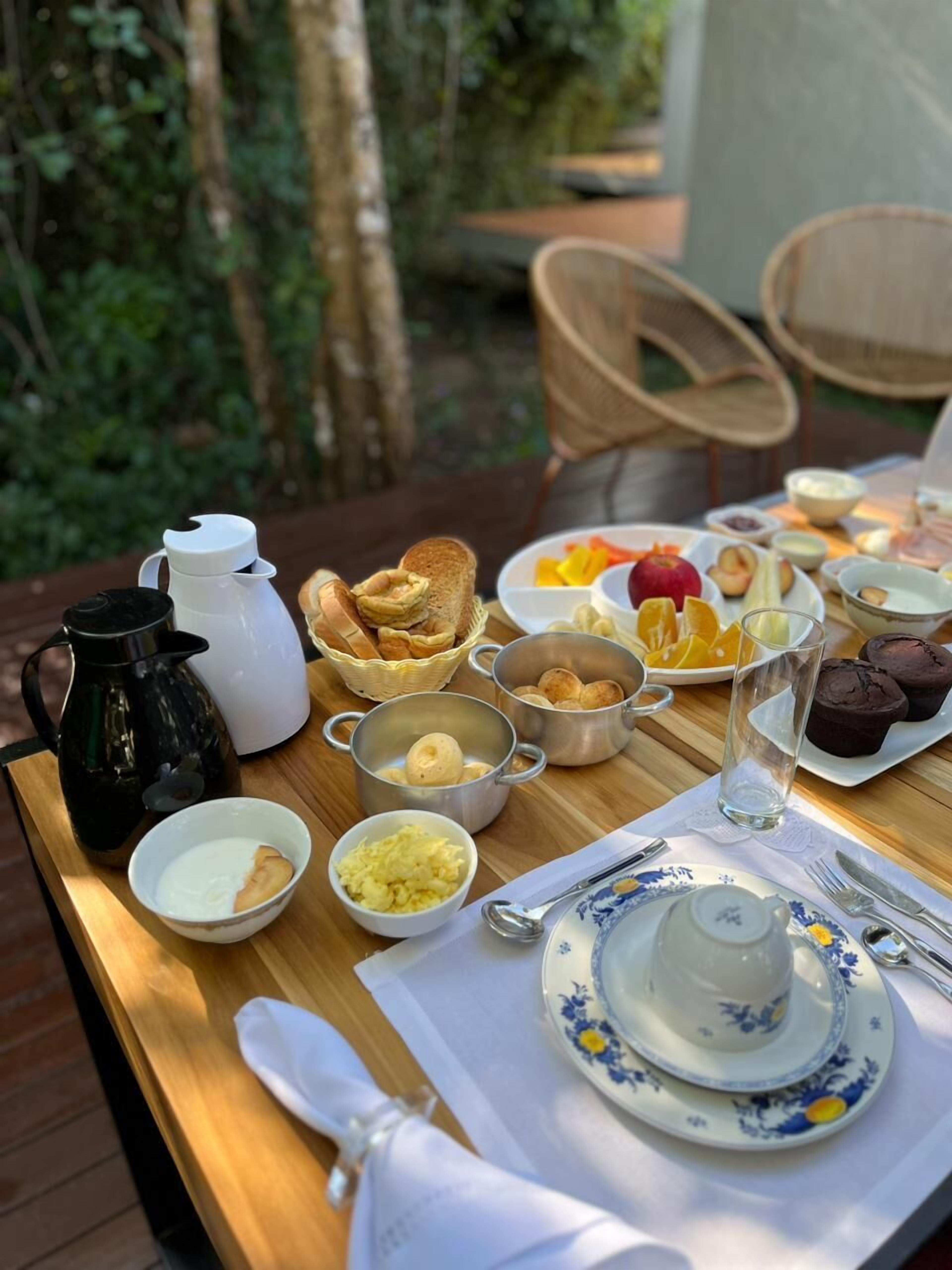 Mesa de café da manhã montada ao ar livre, com diversos alimentos, um bule e um jarro.