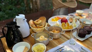 Mesa de café da manhã montada ao ar livre, com diversos alimentos, um bule e um jarro.