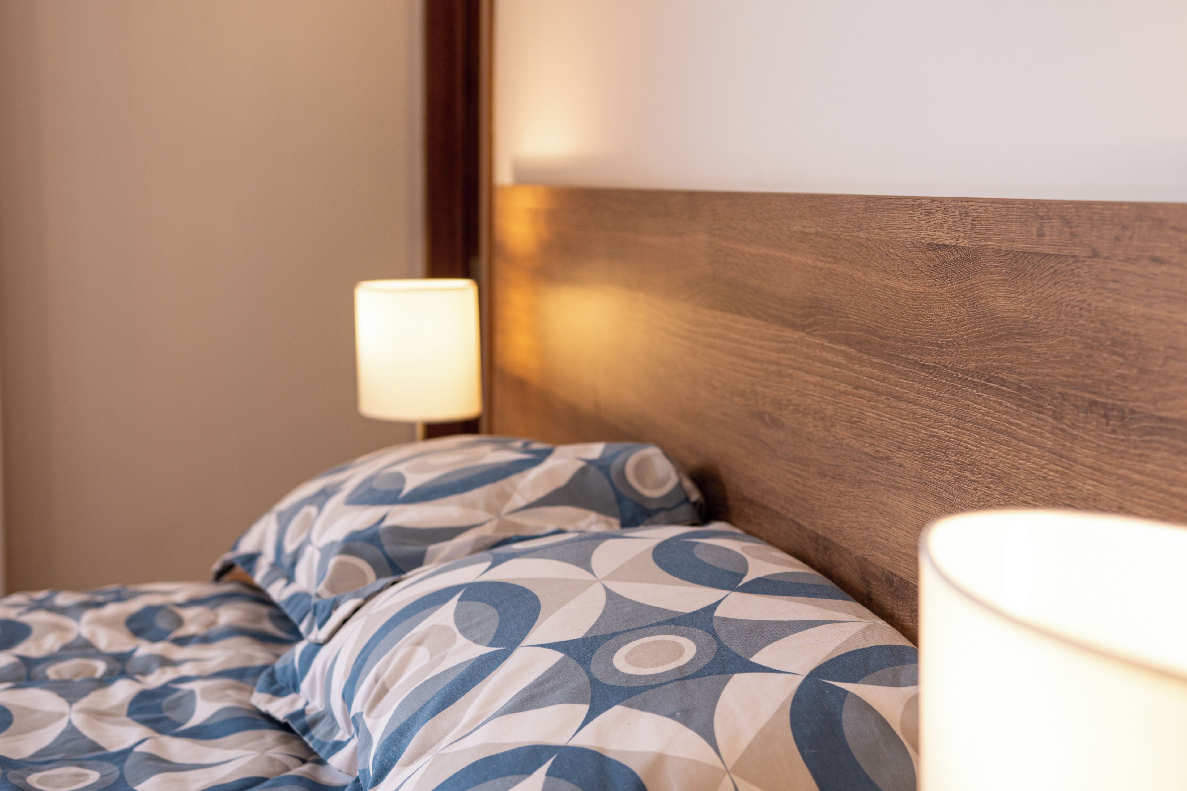 Uma cama bem arrumada com roupa de cama estampada, um abajur e uma cabeceira de madeira.