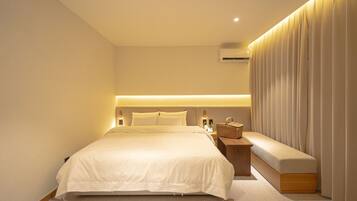 Quarto de hotel moderno com uma cama grande, mesas de cabeceira, um banco e um ar condicionado fixado na parede.