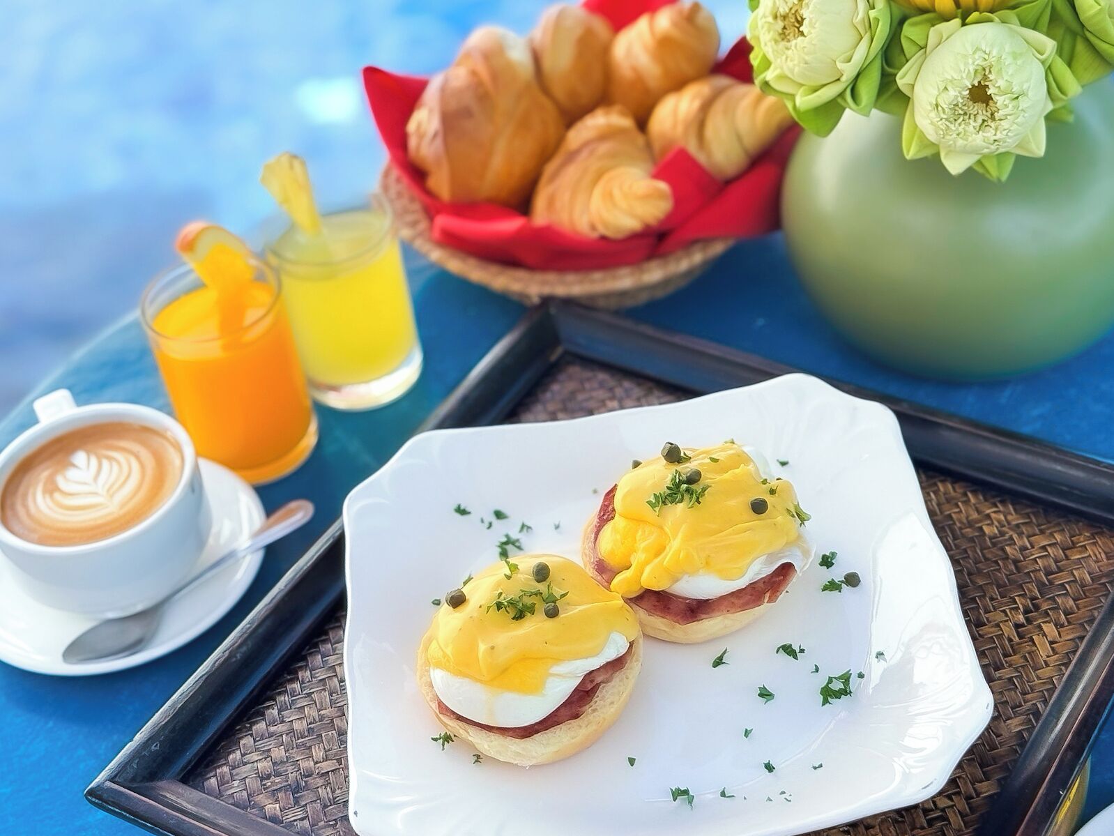 Uma bandeja de café da manhã com dois sanduíches eggs benedict, uma xícara de café, um copo de suco de laranja, uma cesta de croissants e um vaso com flores.