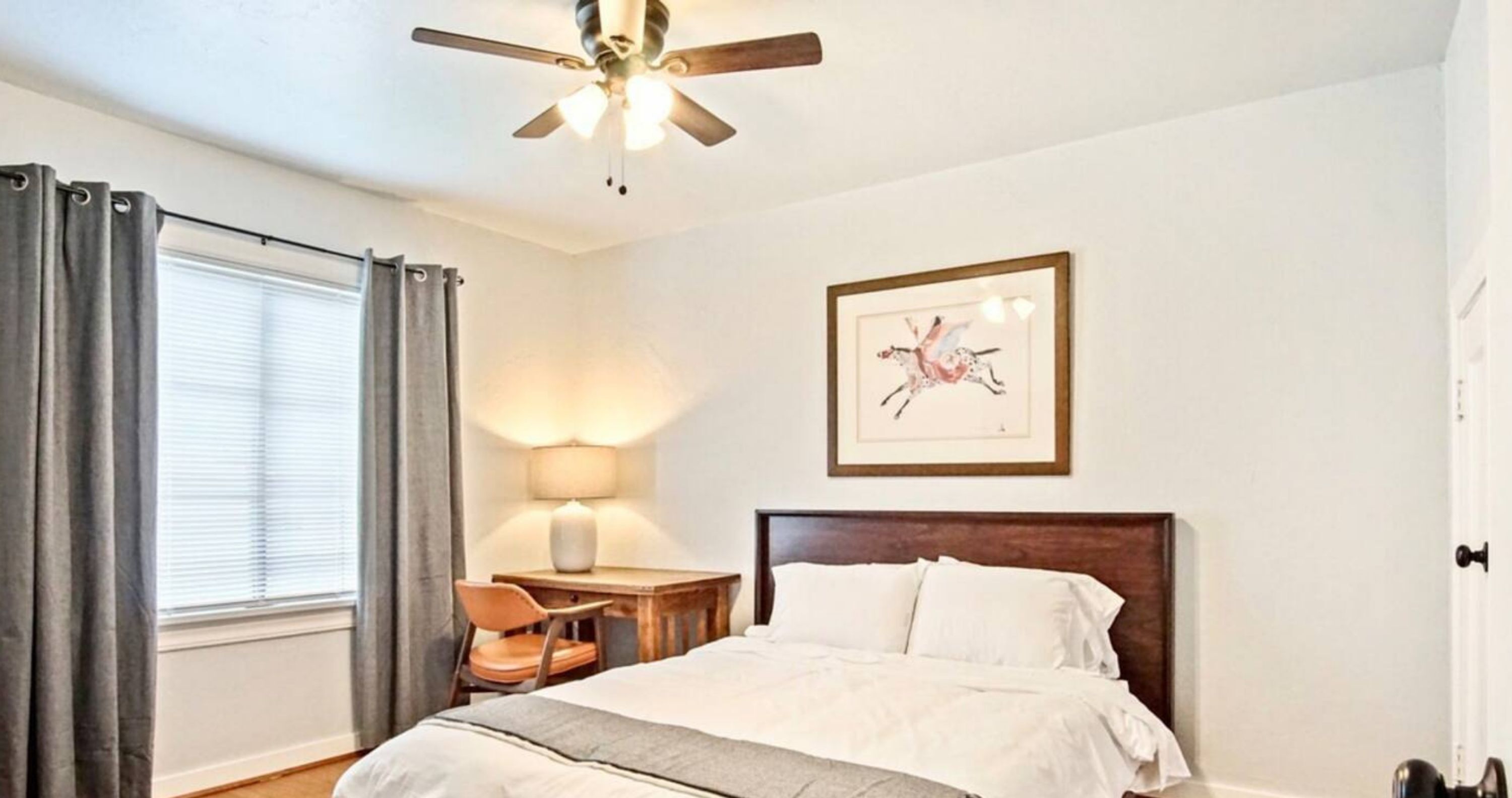 Quarto com cama, escrivaninha, cadeira, ventilador de teto e quadro na parede.