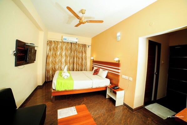 Quarto de hotel com cama, escrivaninha, cadeira, ventoinha no teto e televisão na parede.