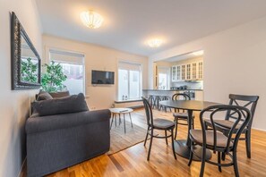 Uma sala de estar moderna com sofá, mesa de jantar e cadeiras, cozinha ao fundo e televisão na parede.