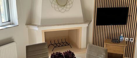 Uma sala de estar moderna com uma mesa de jantar de vidro, duas cadeiras, um vaso com flores, lareira, televisão e painel de parede de madeira.