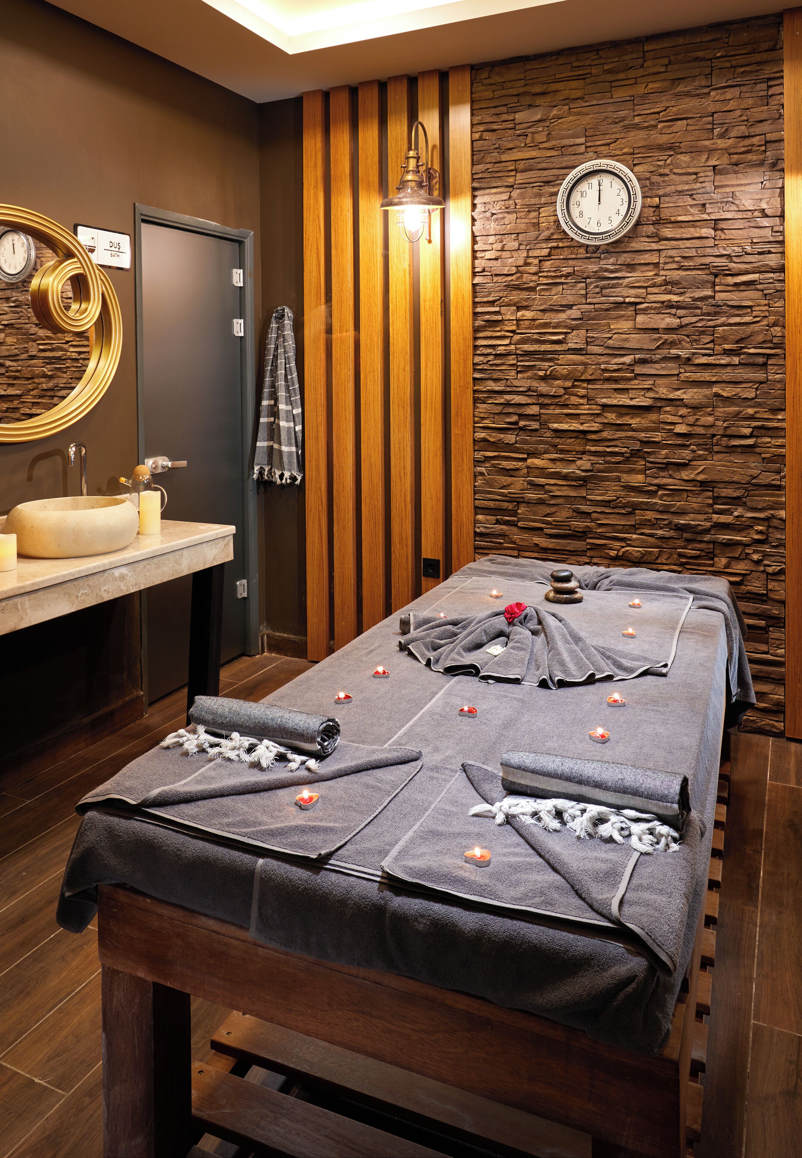 Um espaço de spa com parede de pedra, um banco de madeira, mesa de massagem, pia e relógio.