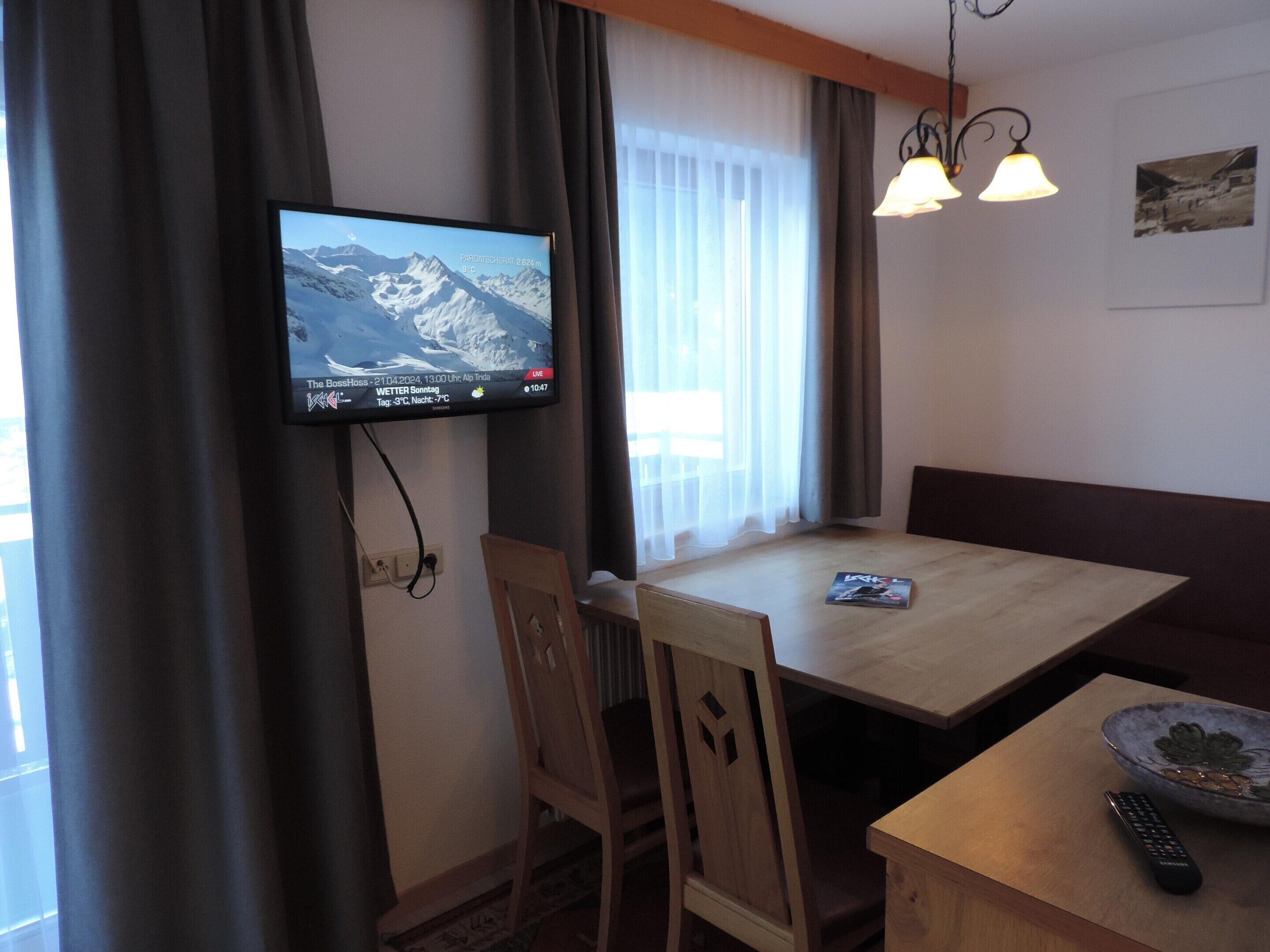 Quarto de hotel com televisão fixada na parede, uma mesa de jantar com cadeiras, um controle remoto e uma tigela decorativa sobre a mesa.