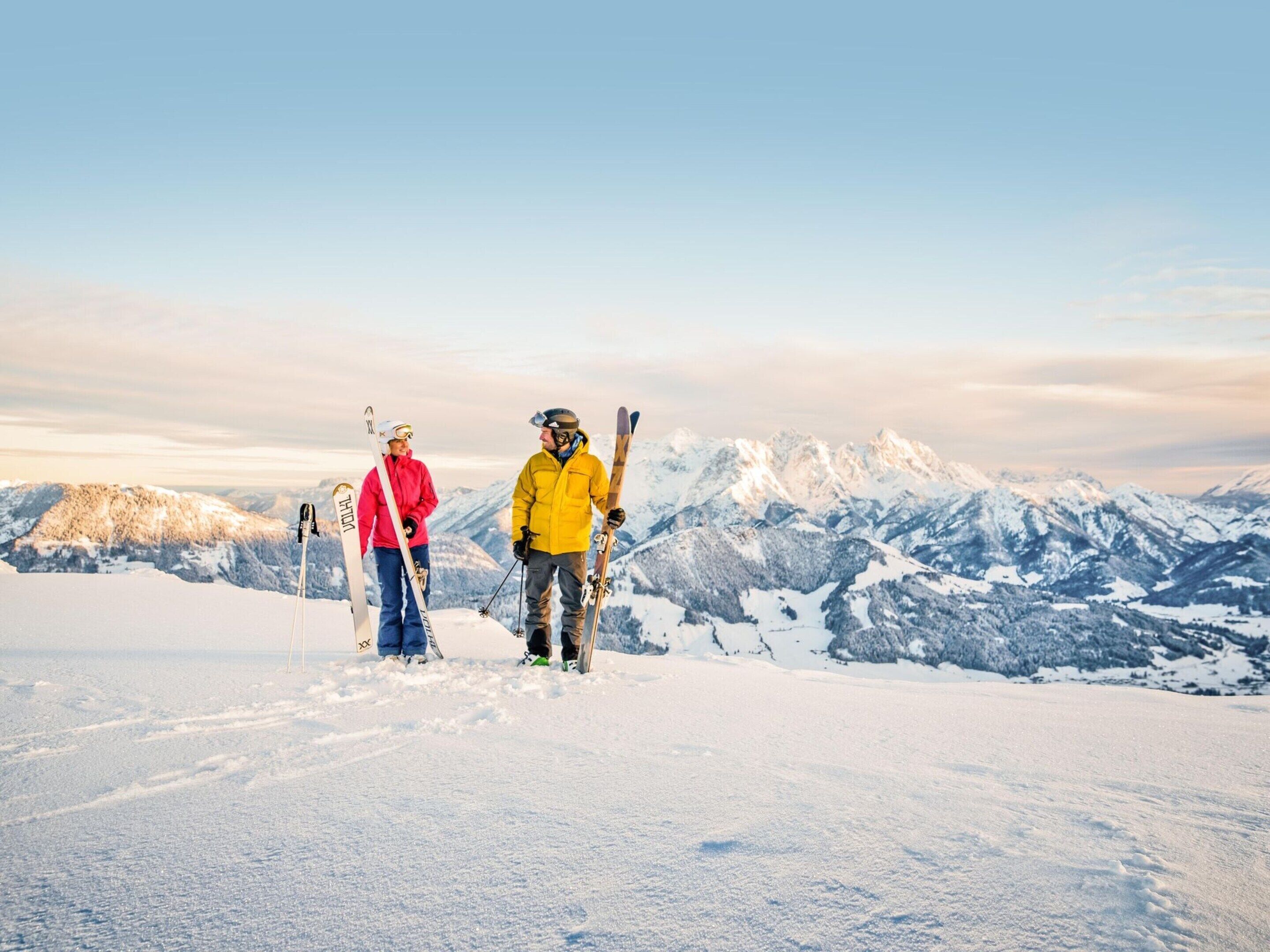 Duas pessoas em pé numa montanha nevada com esquis, com vista para uma cadeia de montanhas cobertas de neve.