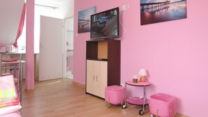 Quarto com decoração em tons de rosa, incluindo televisão, pufes rosa, uma pequena mesa com luminária e obras de arte nas paredes.