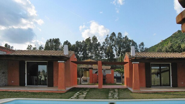 Uma área à beira da piscina com vista desobstruída para uma piscina, dois edifícios com telhados de barro vermelho e um gramado.