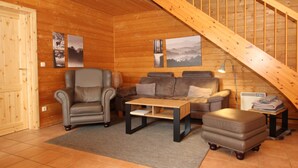 Interior acolhedor de uma cabana de madeira com sofá, poltrona, banquinho e mesa de centro. Uma escada leva a um andar superior.