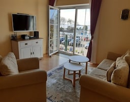 Uma sala de estar com dois sofás, uma mesinha, uma TV e uma porta de vidro deslizante que dá para uma varanda com vista para carros estacionados e árvores.
