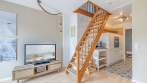 Uma sala de estar moderna com escada de madeira, TV de tela plana em suporte de madeira e área de cozinha ao fundo.