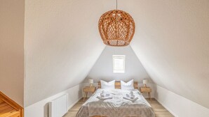 Quarto com teto inclinado, cama com roupa de cama branca, duas mesas de cabeceira com abajures e um banco de madeira aos pés da cama.