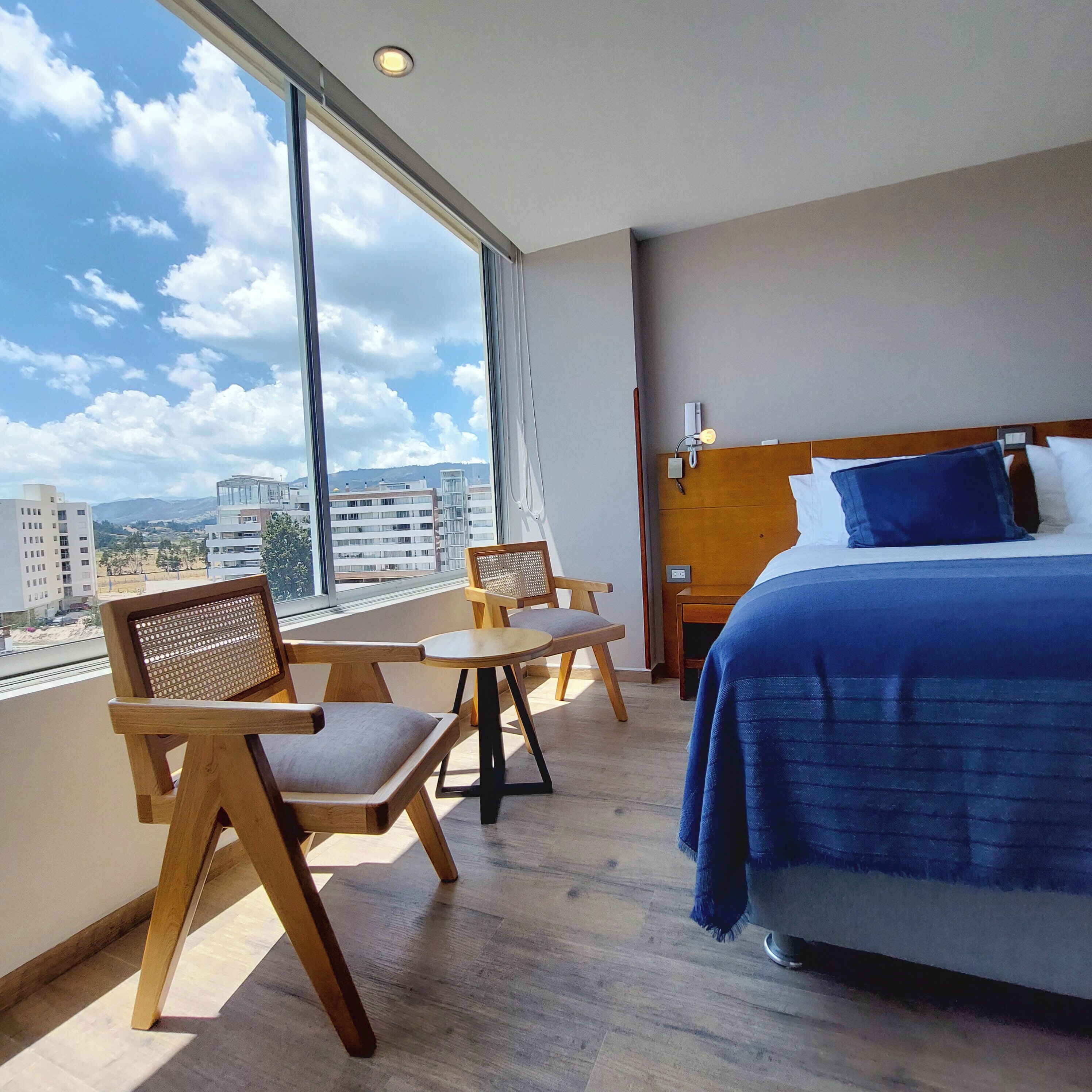 Quarto de hotel com uma janela grande, cama com manta azul, cadeiras de madeira, mesa pequena e mesa de cabeceira com abajur.