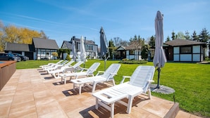 Uma área à beira da piscina com espreguiçadeiras e guarda-sóis, um gramado e edifícios ao fundo.