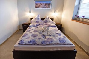 Cama bem arrumada com colcha estampada e almofadas, duas mesas de cabeceira com luminárias, um quadro na parede e uma janela com persianas.