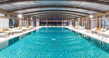 Uma piscina interior com chão ladrilhado, espreguiçadeiras e teto de vidro.