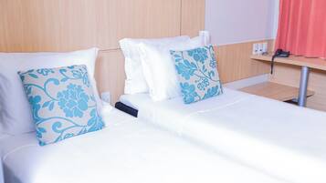 Quarto de hotel com duas camas, cada uma com roupa de cama branca e almofadas florais azuis. Há uma cabeceira de madeira, uma mesinha com um telefone e uma cortina vermelha.