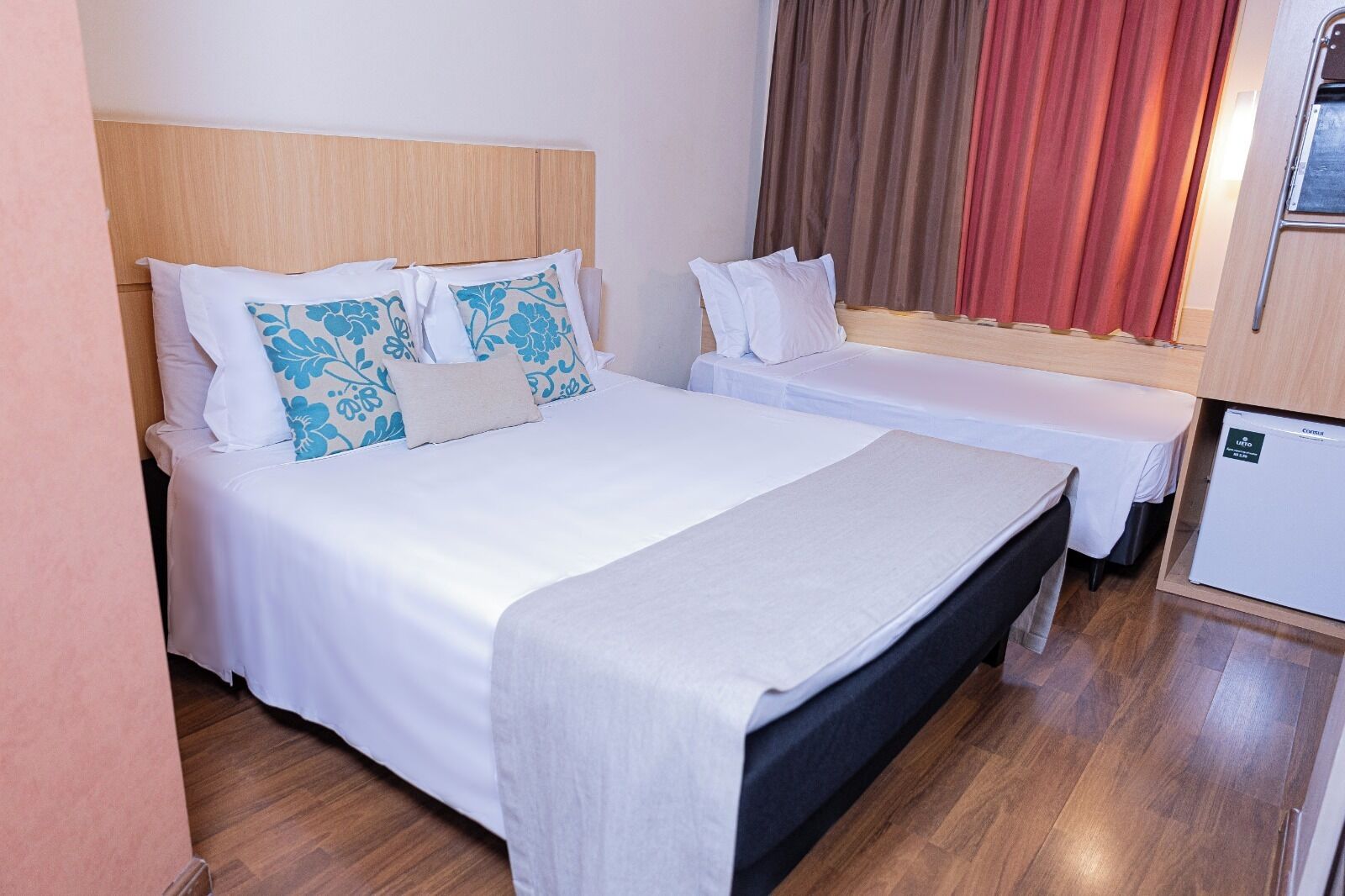 Cama de casal com cabeceira de madeira, roupa de cama branca e almofadas decorativas. Uma pequena mesa de cabeceira e um frigorífico são visíveis.