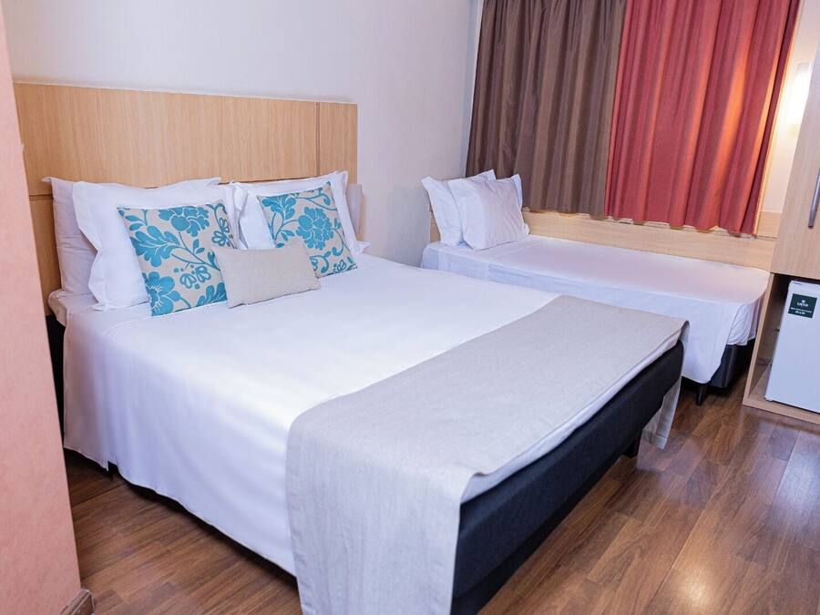 Cama de casal com cabeceira de madeira, roupa de cama branca e almofadas decorativas. Uma pequena mesa de cabeceira e um frigorífico são visíveis.