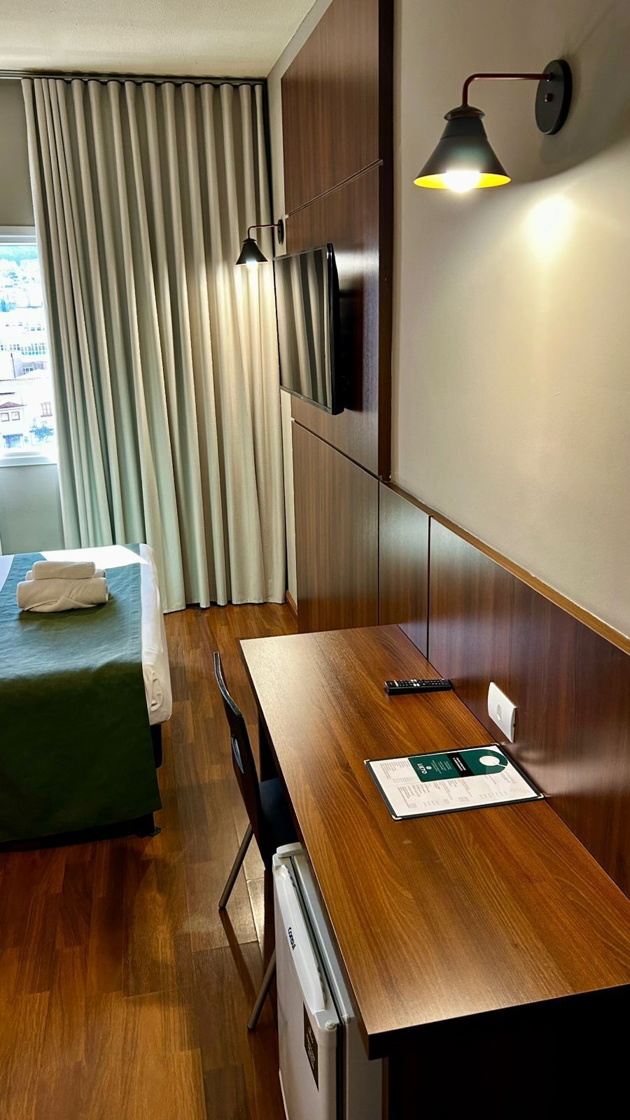 Quarto de hotel com secretária de madeira, cama com roupa de cama verde, televisão presa à parede e janela com cortinas.