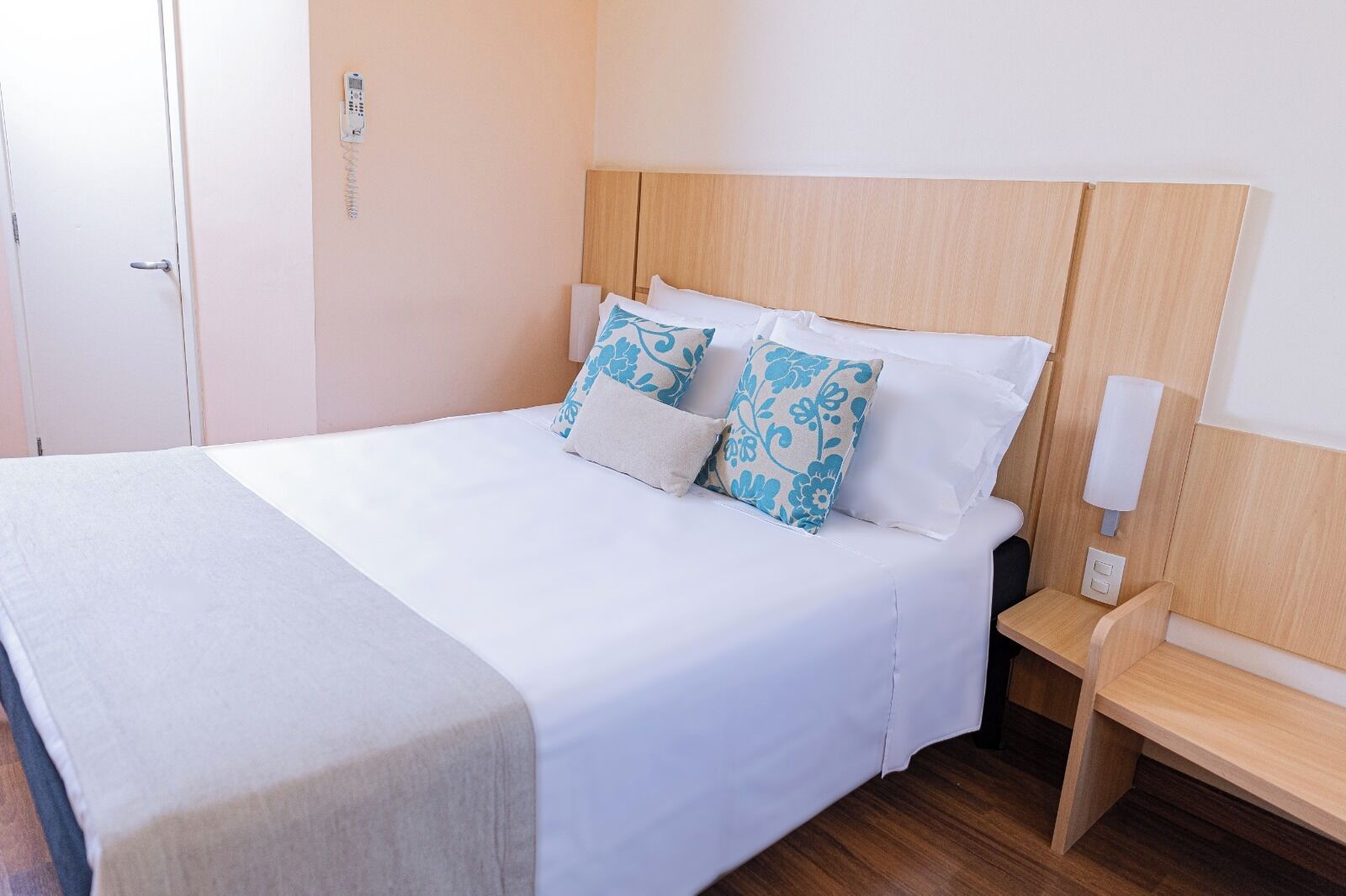Uma cama bem arrumada com roupa de cama branca e almofadas estampadas com flores, uma mesa de cabeceira de madeira com abajur e piso de madeira.
