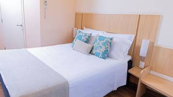 Uma cama bem arrumada com roupa de cama branca e almofadas estampadas com flores, uma mesa de cabeceira de madeira com abajur e piso de madeira.