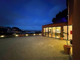 Terraço no telhado com vista para a cidade ao entardecer, com área de bar, portas de vidro e plantas em vasos.