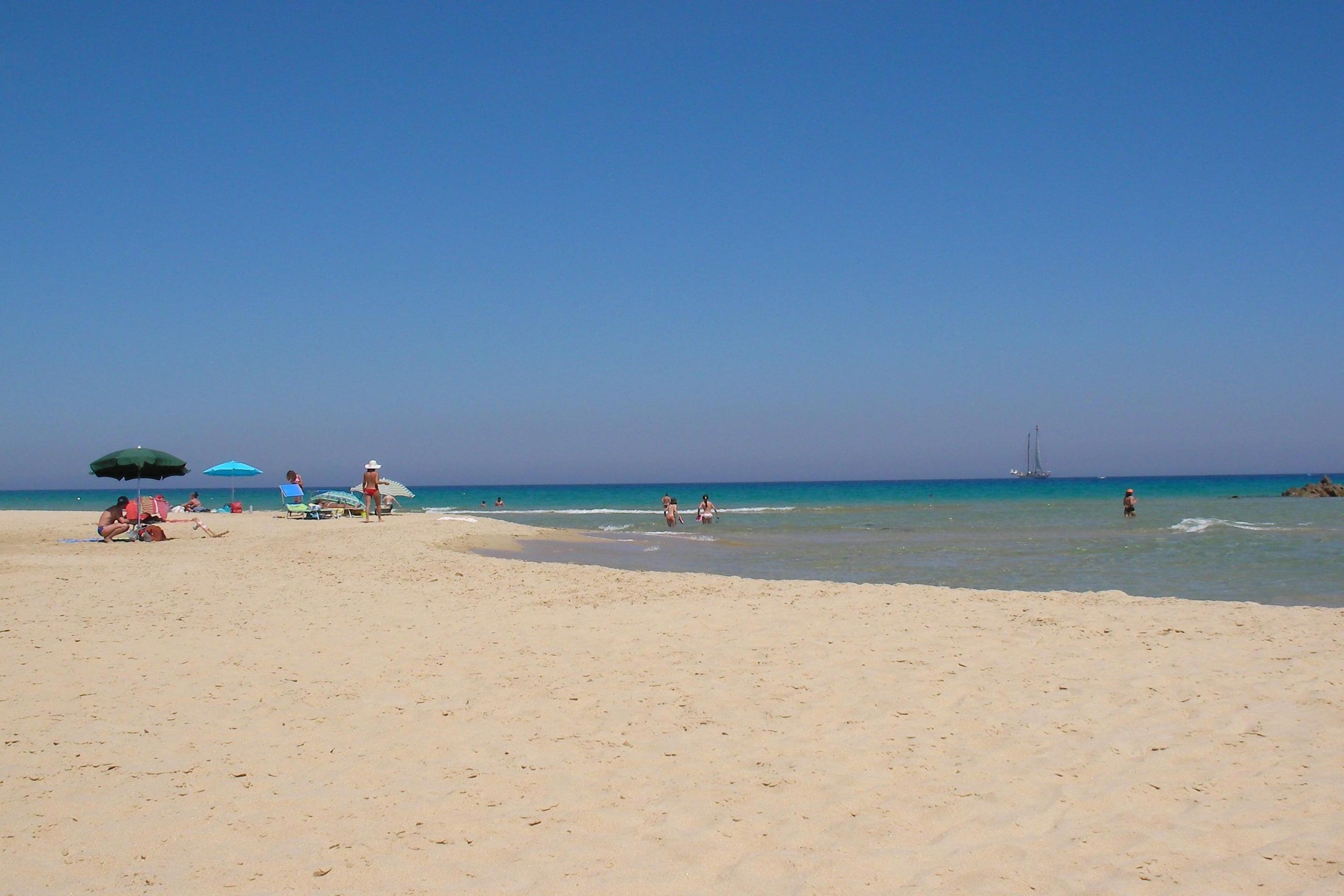 Uma praia com céu claro, areal arenoso e algumas pessoas e guarda-sóis.