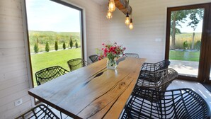 Uma sala de jantar com uma mesa de madeira, cadeiras pretas e um vaso com flores. Grandes janelas oferecem vista para um campo gramado.