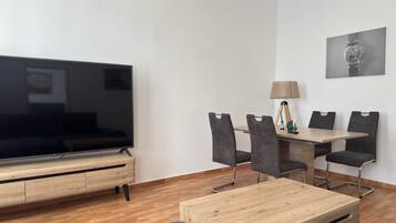 Uma sala de estar moderna com televisão de tela plana, mesa de centro de madeira, sofás cinzas e um espaço de jantar com mesa e cadeiras.