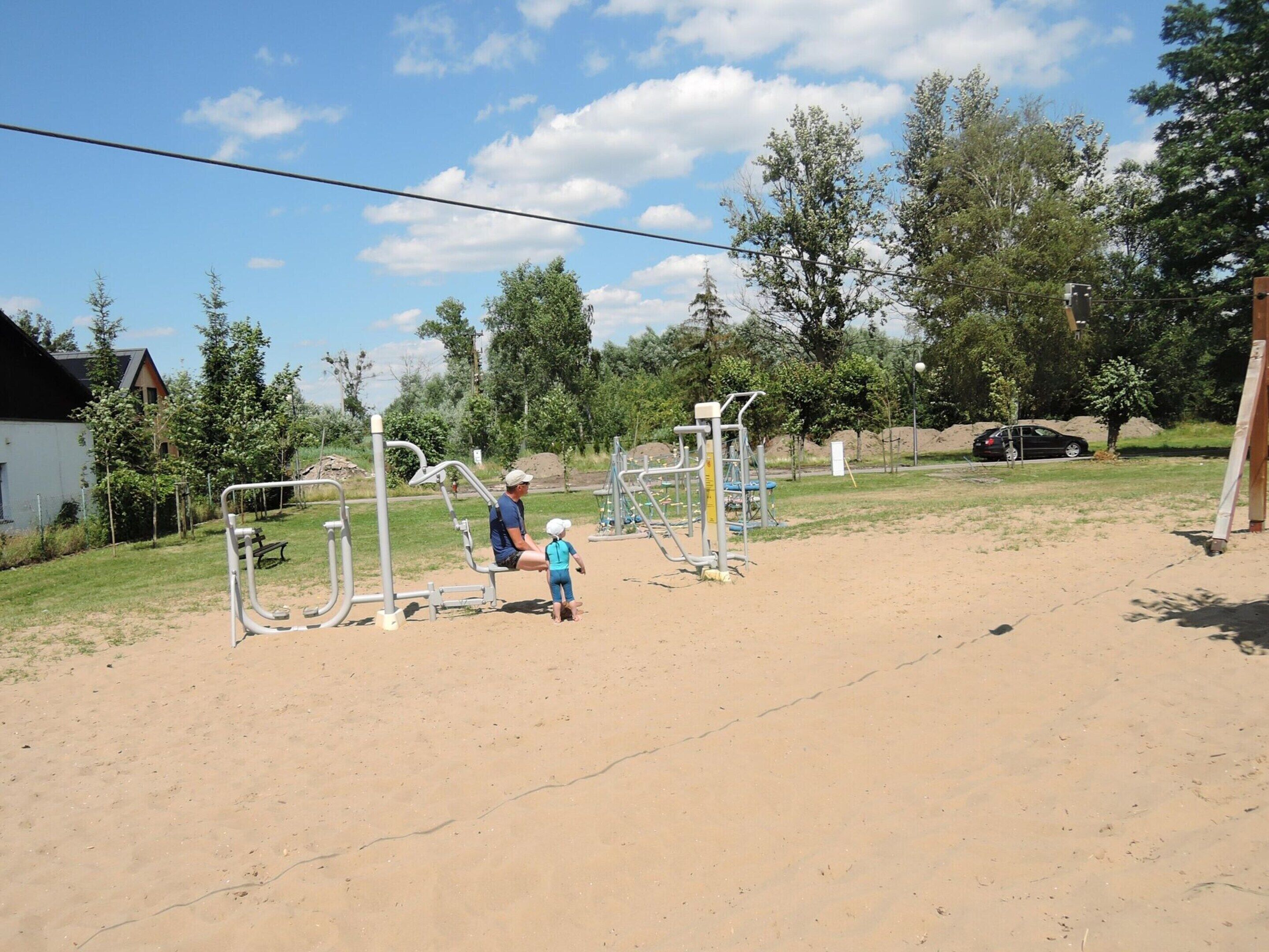 Academia ao ar livre com equipamentos de exercício, chão de terra e árvores ao fundo.