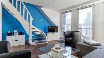 Uma sala de estar moderna com uma parede azul de destaque, uma escada branca, uma televisão de tela plana, uma mesa de centro de vidro e uma varanda com vista.