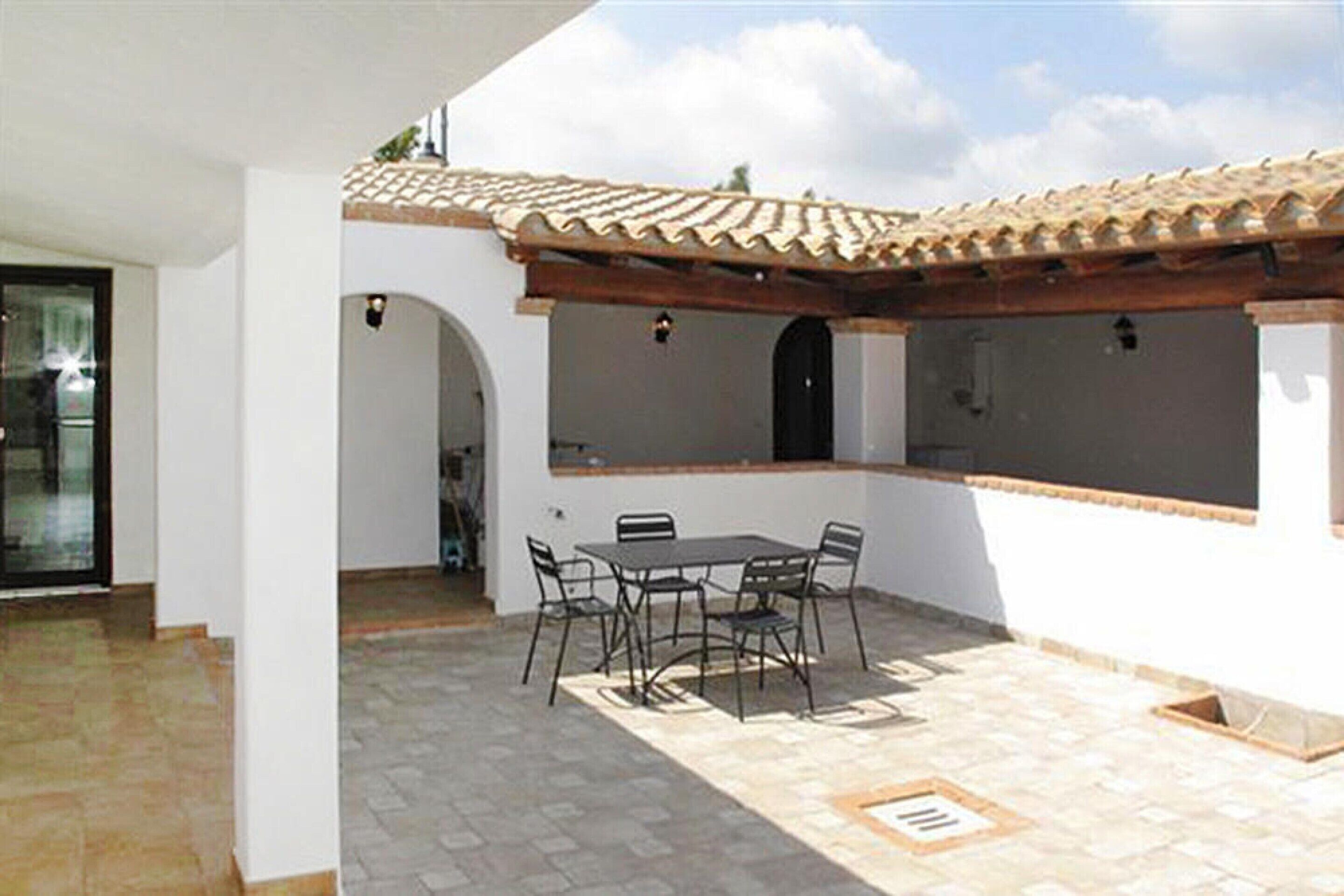 Uma área de pátio externa com telhado de azulejos, mesa e cadeiras, e uma parede branca.