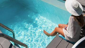 Uma pessoa sentada à beira de uma piscina com água cristalina, usando um chapéu branco e uma blusa de cor clara.