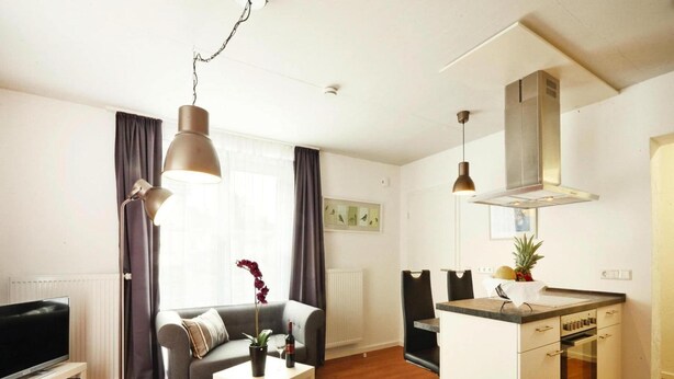 Uma sala de estar moderna com um sofá, um banquinho vermelho, uma mesa de centro e uma TV. Há uma cozinha com fogão, forno e uma mesa de jantar com cadeiras.