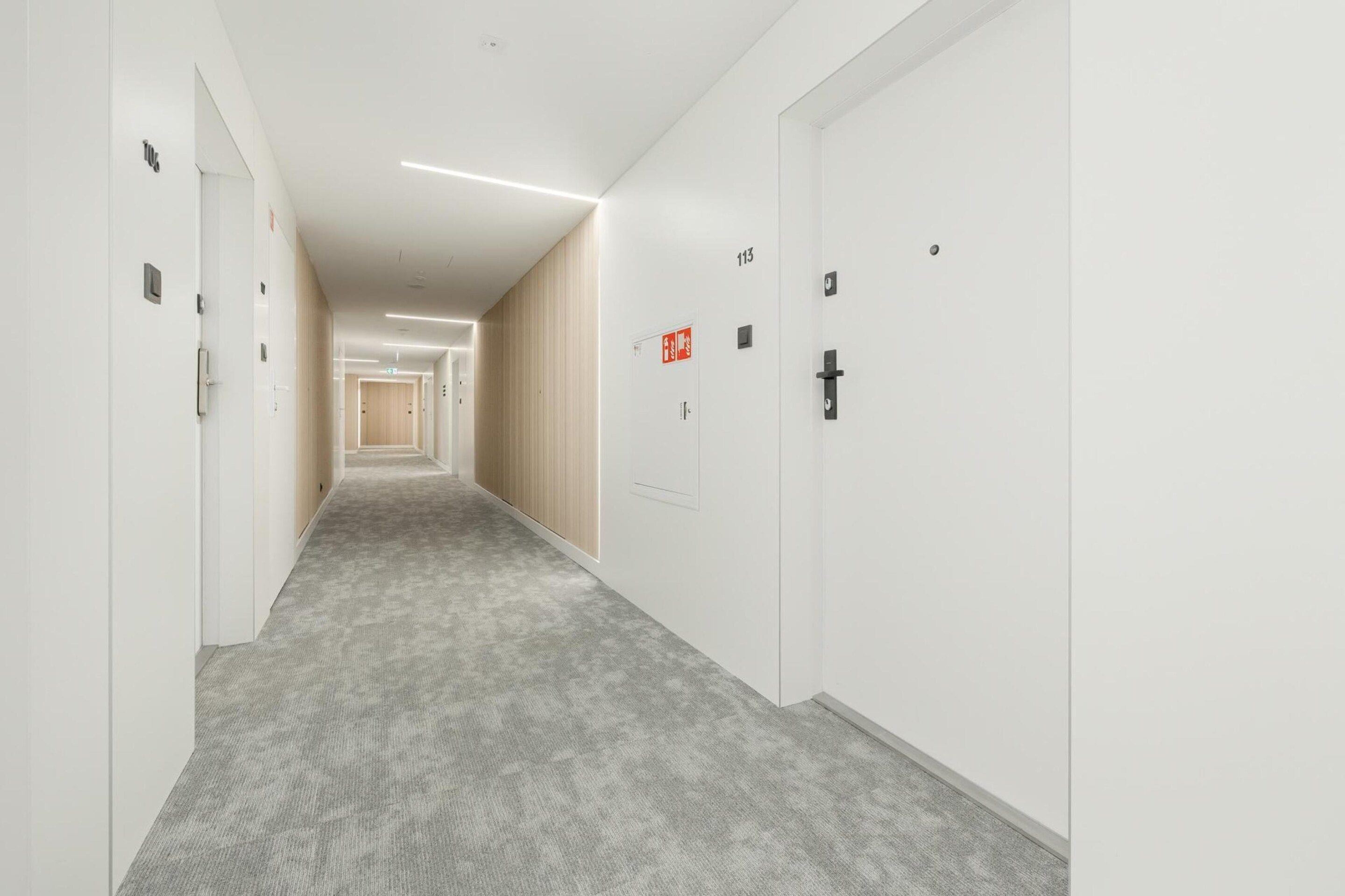 Corredor comprido e estreito com piso de carpete e paredes brancas, que leva a várias portas com números de quarto.
