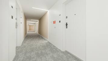Corredor comprido e estreito com piso de carpete e paredes brancas, que leva a várias portas com números de quarto.